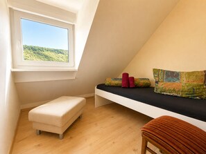 Um quarto pequeno e acolhedor com teto inclinado, uma janela com vista para uma paisagem verde, um banco com almofadas e piso de madeira.