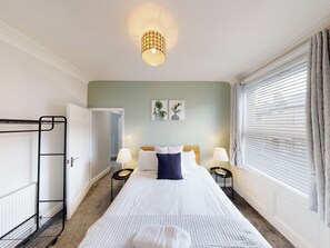 Quarto de hotel com cama, duas luminárias de cabeceira, iluminação embutida na parede, janela com persianas e porta que dá para outro cômodo.