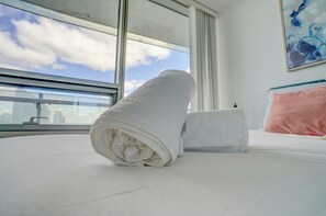 Uma cama bem arrumada com uma toalha enrolada e um travesseiro, uma janela com vista para a cidade e um quadro na parede.