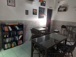 Quarto com mesa de vidro, cadeiras de vime, estante de livros e um pequeno bar.