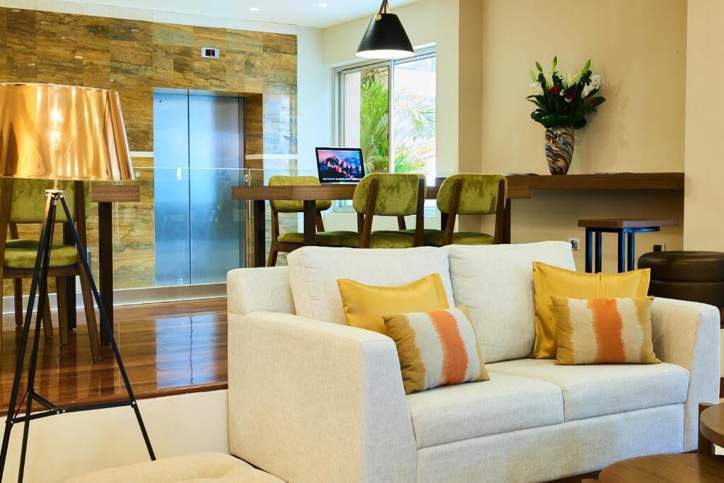 Um lobby moderno com um sofá, cadeiras, uma luminária, um laptop sobre uma mesa e uma planta.
