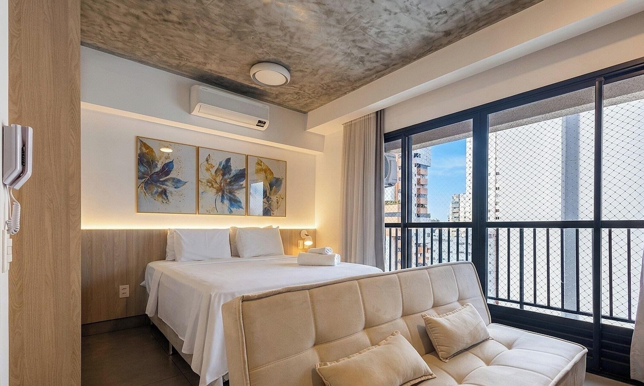 Quarto de hotel com uma cama, um sofá, uma janela grande com vista e obras de arte na parede.