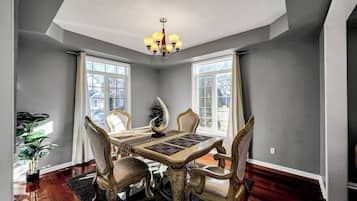 Uma sala de jantar com um candelabro, uma mesa com quatro cadeiras e janelas amplas.