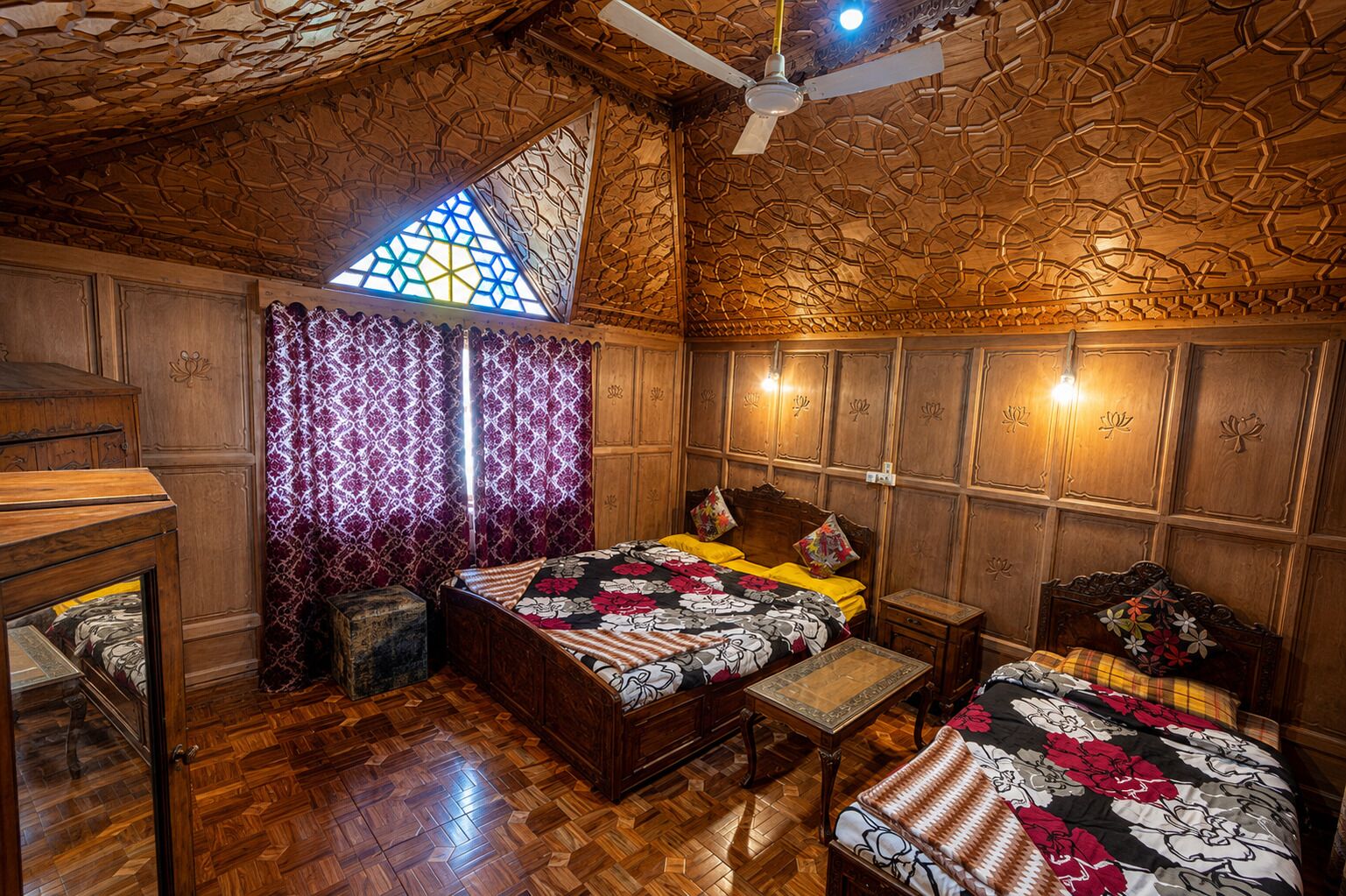 Quarto com teto de madeira, cama com roupa de cama floral, uma mesa de madeira e uma janela com cortinas estampadas.