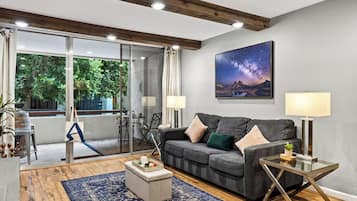 Uma sala de estar moderna com um sofá cinzento, uma mesa de centro de vidro e uma grande janela com vista para o verde.