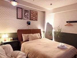 Quarto de hotel com cama, mesa de cabeceira, luminária, obra de arte na parede e planta pendente.