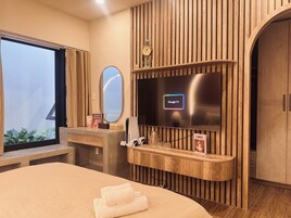 Quarto de hotel com uma cama grande, uma televisão fixada num painel de madeira, uma mesa de cabeceira com uma luminária e uma janela com cortinas.