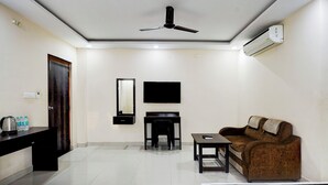 Uma sala de estar com ventilador de teto, televisão de tela plana, sofá, mesa de centro e uma mesinha com chaleira e garrafas de água.