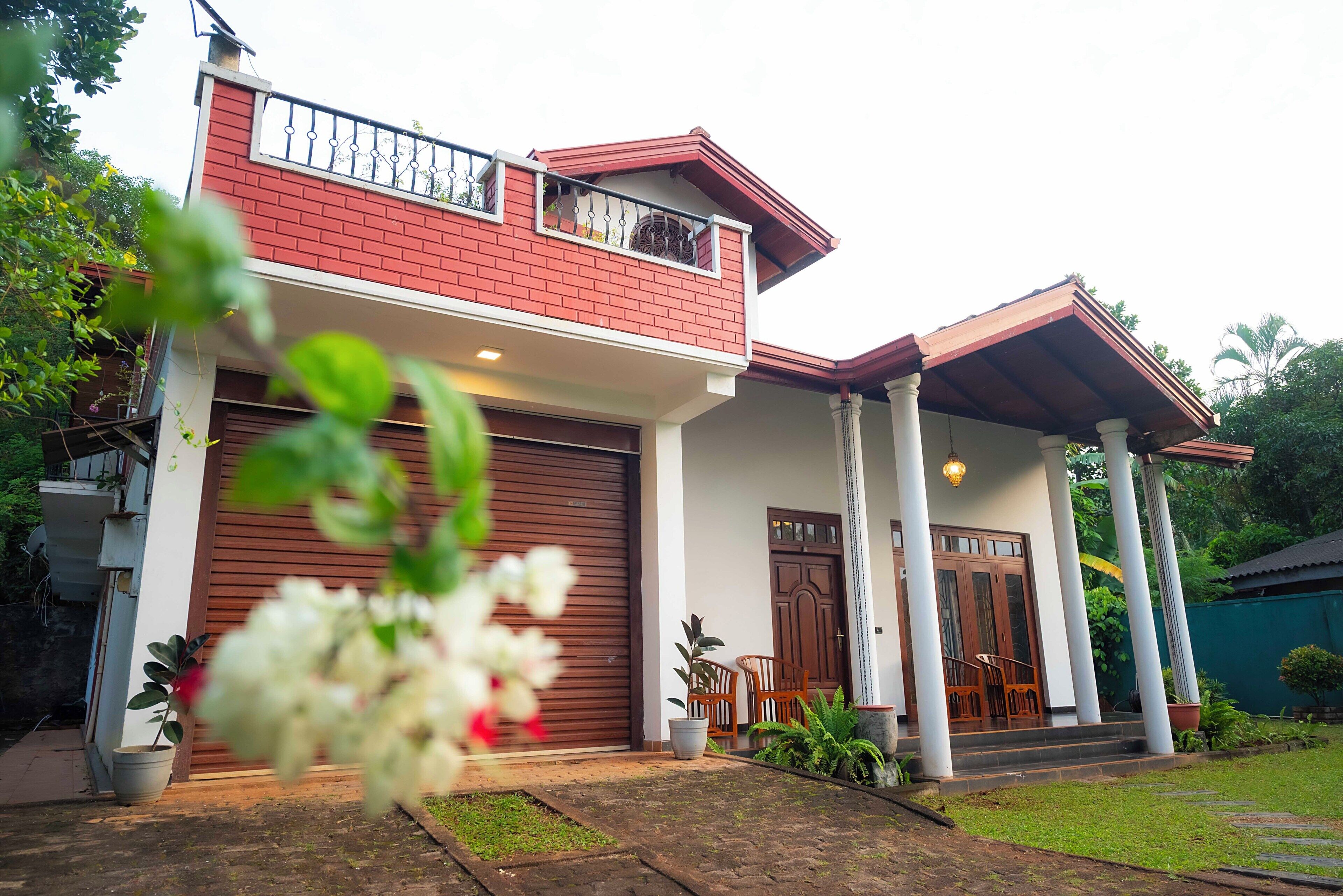 Uma casa de dois andares com telhado vermelho, varanda e um alpendre coberto com colunas.