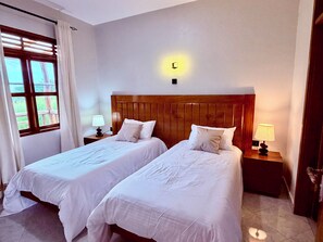 Quarto de hotel com duas camas, janela com cortinas, luminária na parede e mesa de cabeceira com abajur.