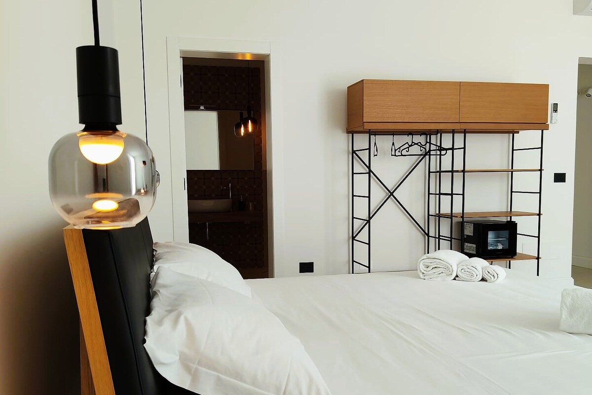 Cama com estrutura de madeira, luminária pendente e televisão fixada na parede.