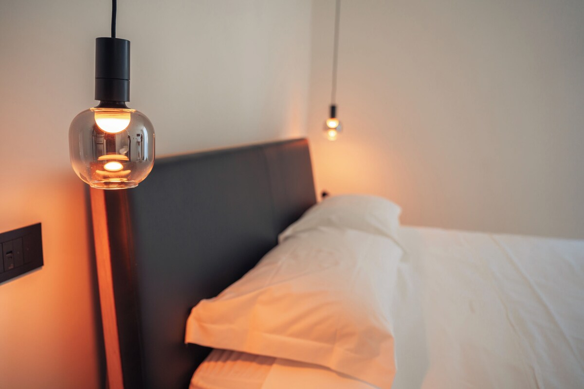 Uma cama com lençóis brancos e cabeceira escura, uma tomada e um lustre.