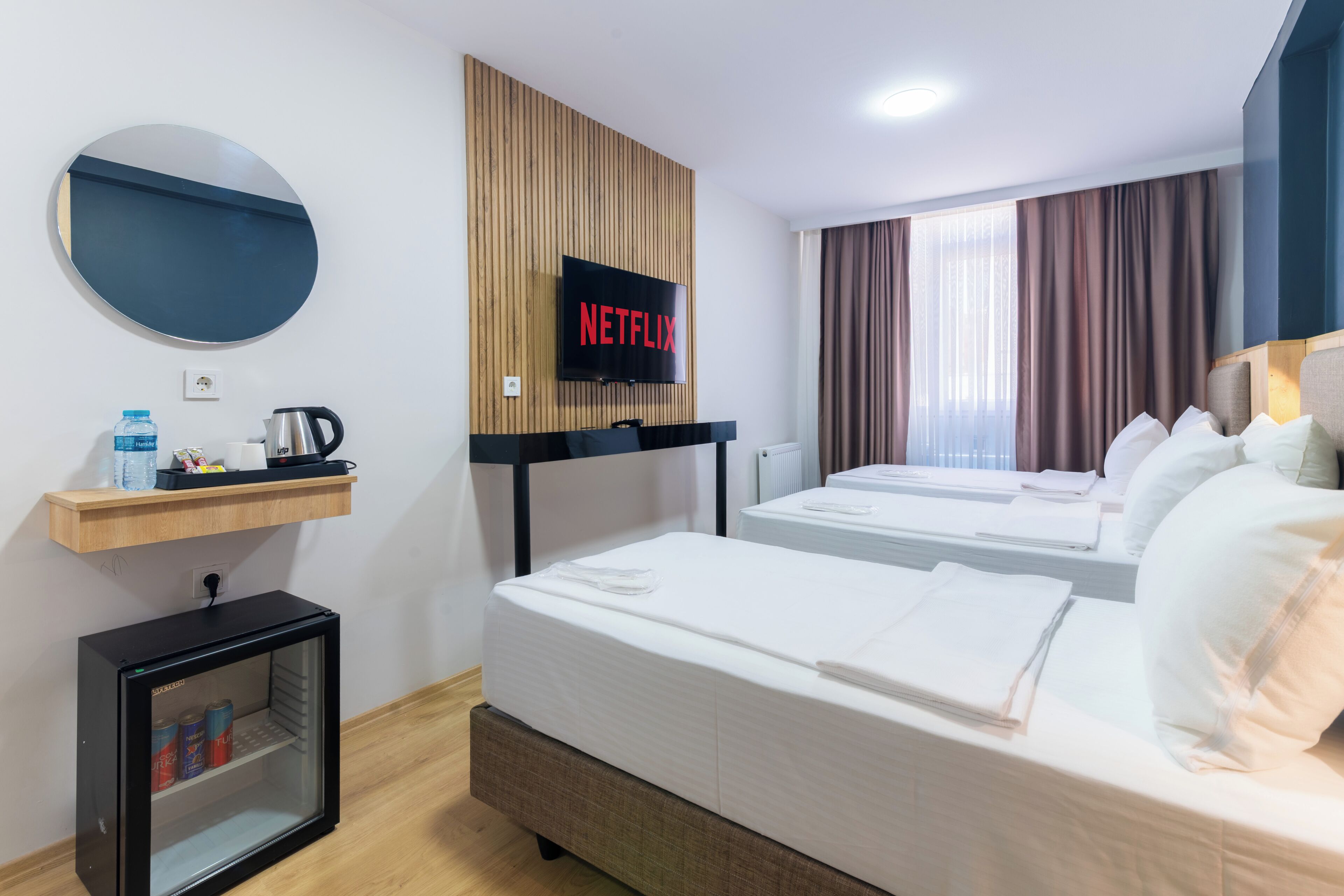 Quarto de hotel com duas camas, televisão montada em um painel de madeira, minibar e janela com cortinas.