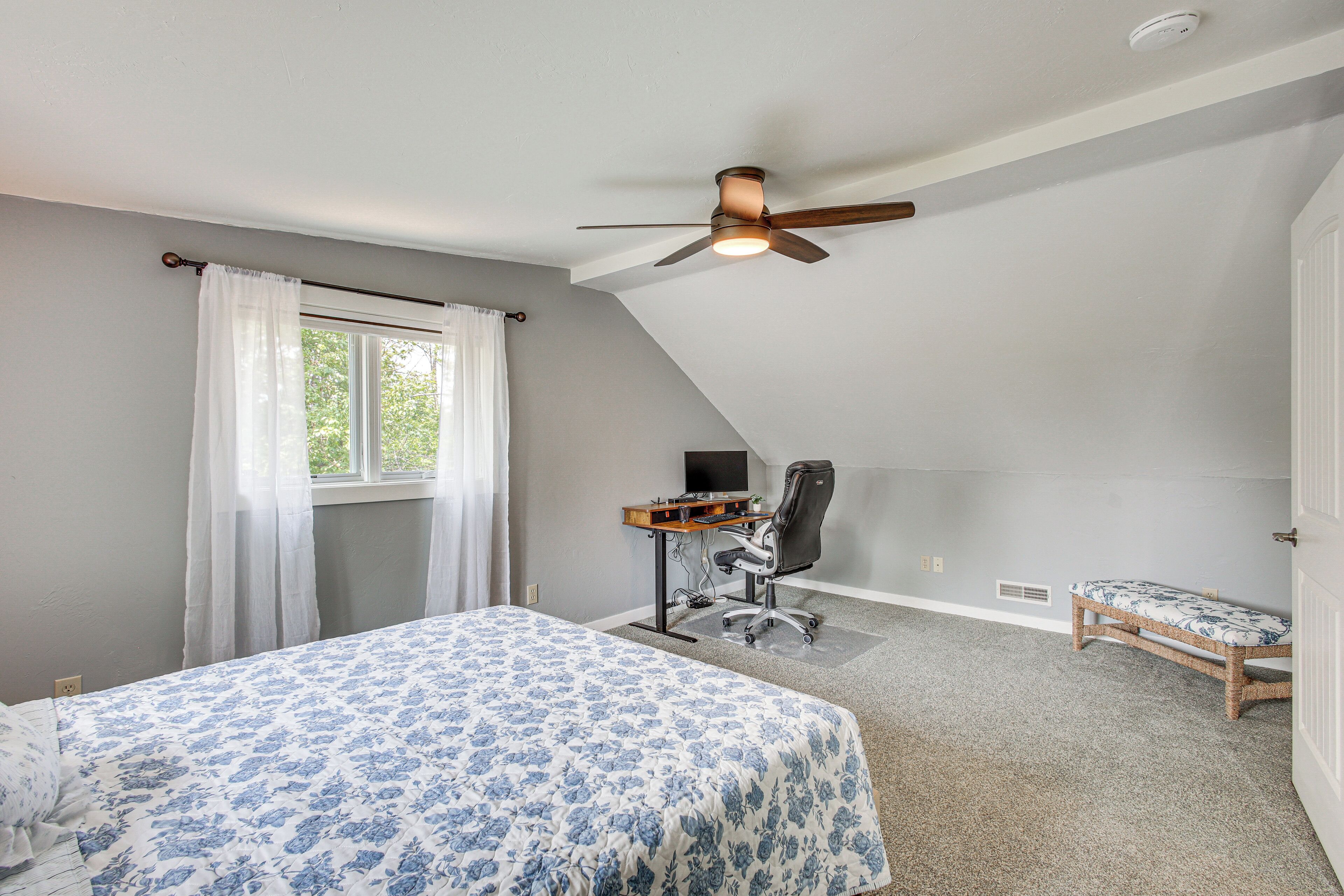 Quarto com cama, escrivaninha, cadeira, ventilador de teto e janela com cortinas.