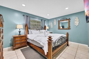 Quarto com cama, mesas de cabeceira, penteadeira, janela com cortinas, espelho e quadros nas paredes.