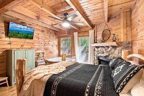 Interior de uma cabana de madeira com uma cama, uma televisão, lareira e janela.