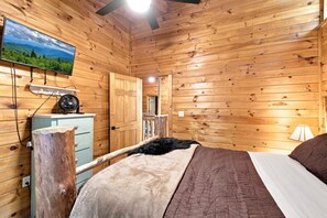 Interior de uma cabana de madeira com uma cama, cômoda, televisão fixada na parede e um ventilador de teto.
