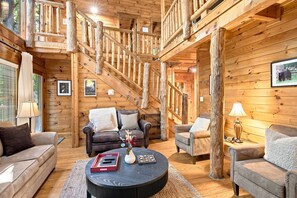 Interior de cabana de madeira com escada, sofás de couro, mesa de centro e lareira.