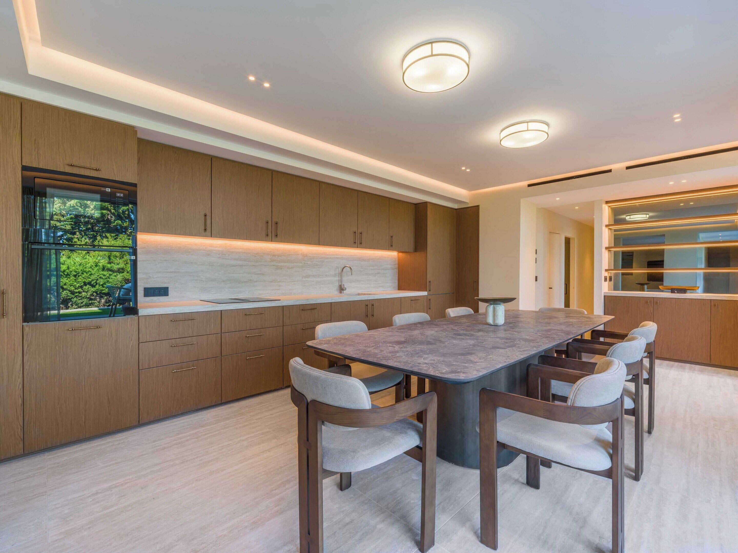 Uma cozinha moderna com eletrodomésticos embutidos, sala de jantar com uma mesa grande e cadeiras, e vista para o verde através de uma janela.