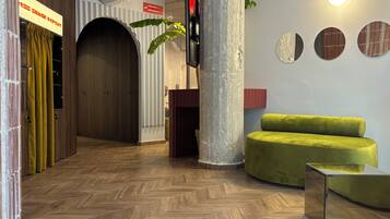 Um hall de hotel com piso de madeira em padrão herringbone, um sofá estofado verde e uma grande porta de madeira.