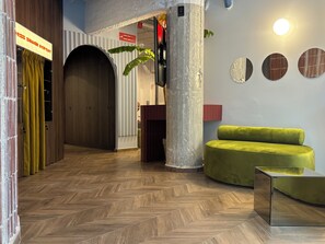 Um hall de hotel com piso de madeira em padrão herringbone, um sofá estofado verde e uma grande porta de madeira.