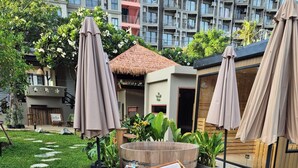 Um terraço com espreguiçadeiras e guarda-sóis, rodeado por vegetação e um edifício ao fundo.