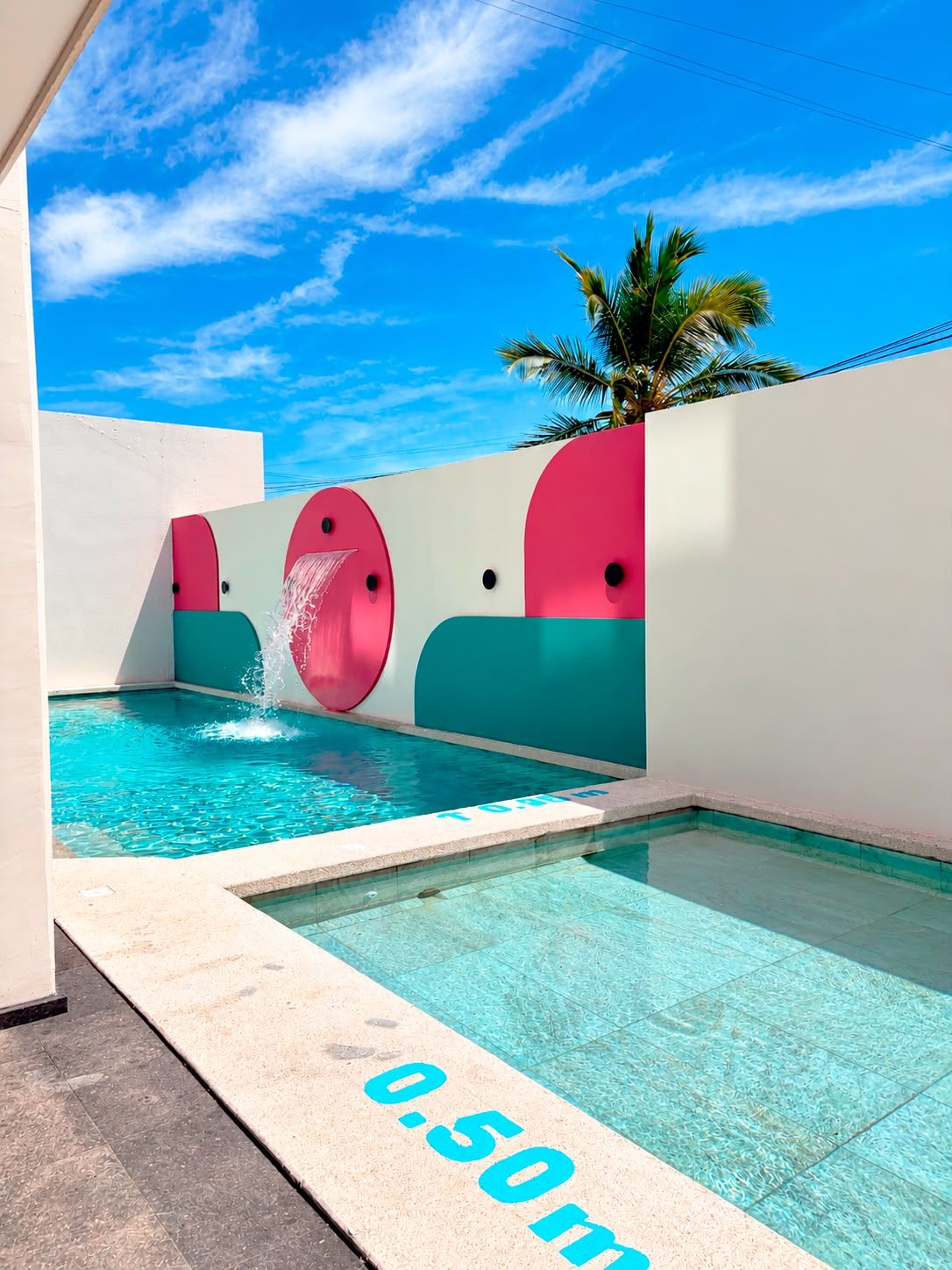 Piscina com superfície de água azul cristalina, rodeada por paredes com formas abstratas e uma palmeira ao fundo.
