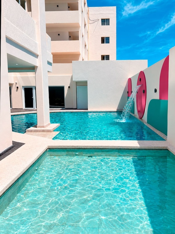 Piscina com água azul cristalina, cercada por um edifício branco com varandas e uma porta.