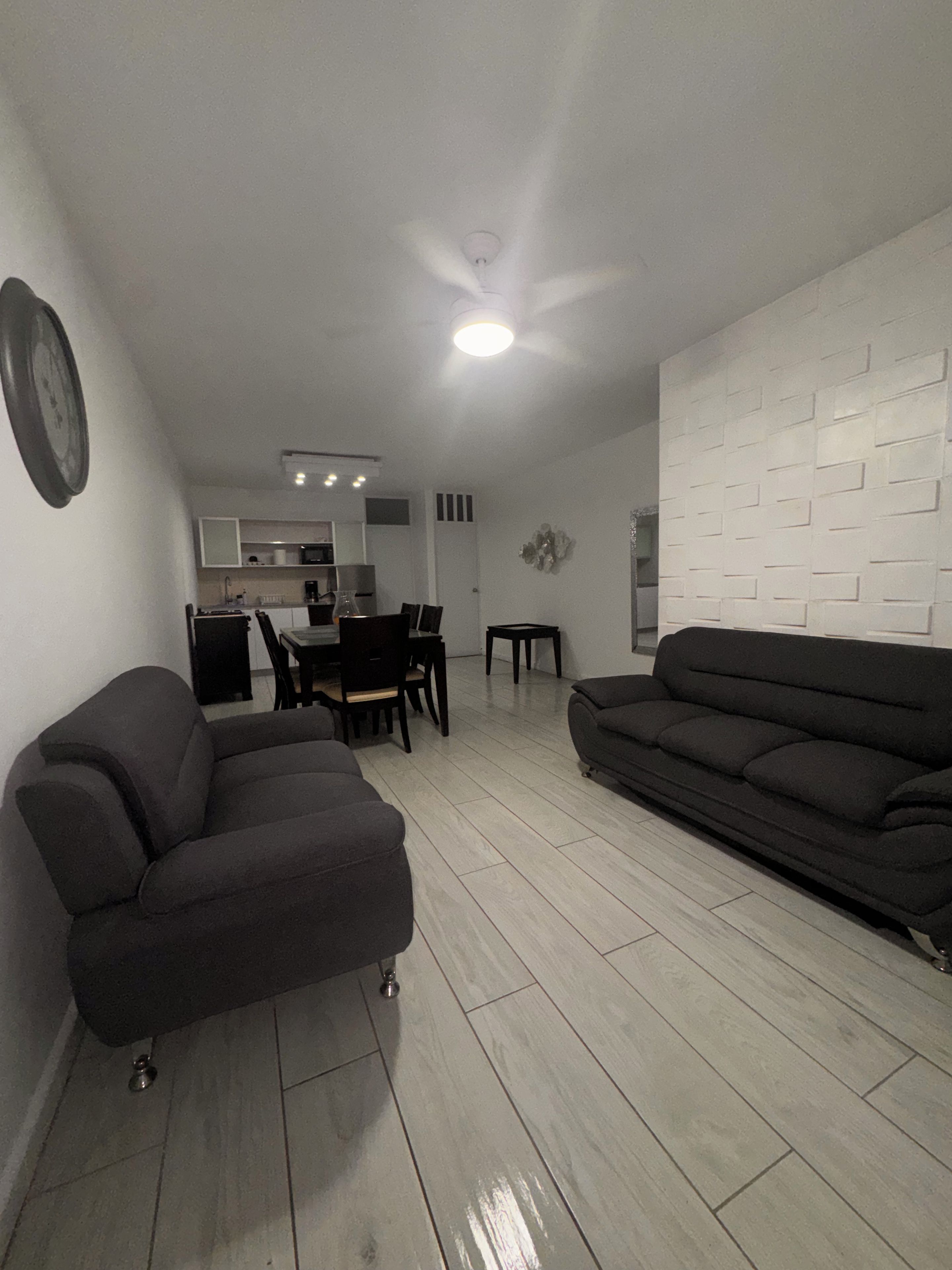 Uma sala de estar moderna com um sofá seccional, uma área de jantar com mesa e cadeiras, e uma cozinha ao fundo.