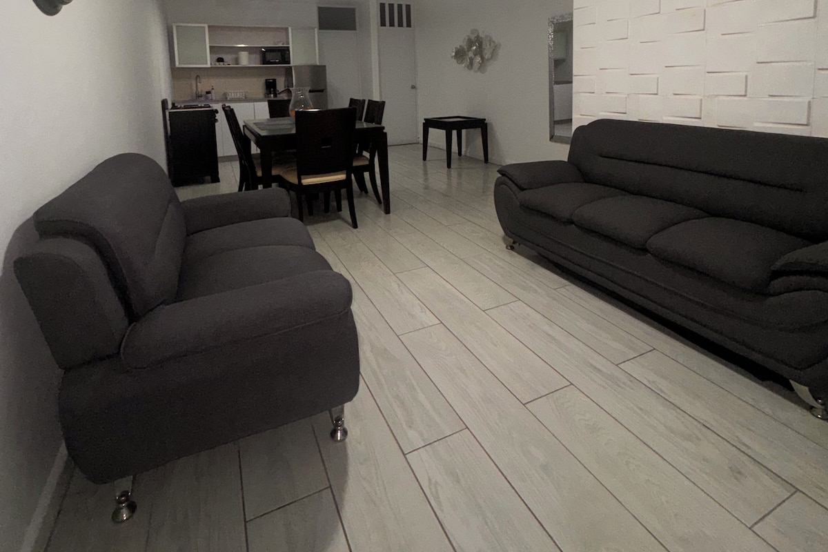 Uma sala de estar moderna com um sofá seccional, uma área de jantar com mesa e cadeiras, e uma cozinha ao fundo.