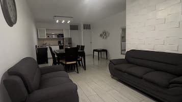 Uma sala de estar moderna com um sofá seccional, uma área de jantar com mesa e cadeiras, e uma cozinha ao fundo.