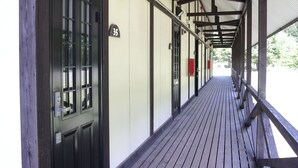 Uma galeria coberta com piso de madeira, grades de metal e portas com números que indicam quartos ou unidades.
