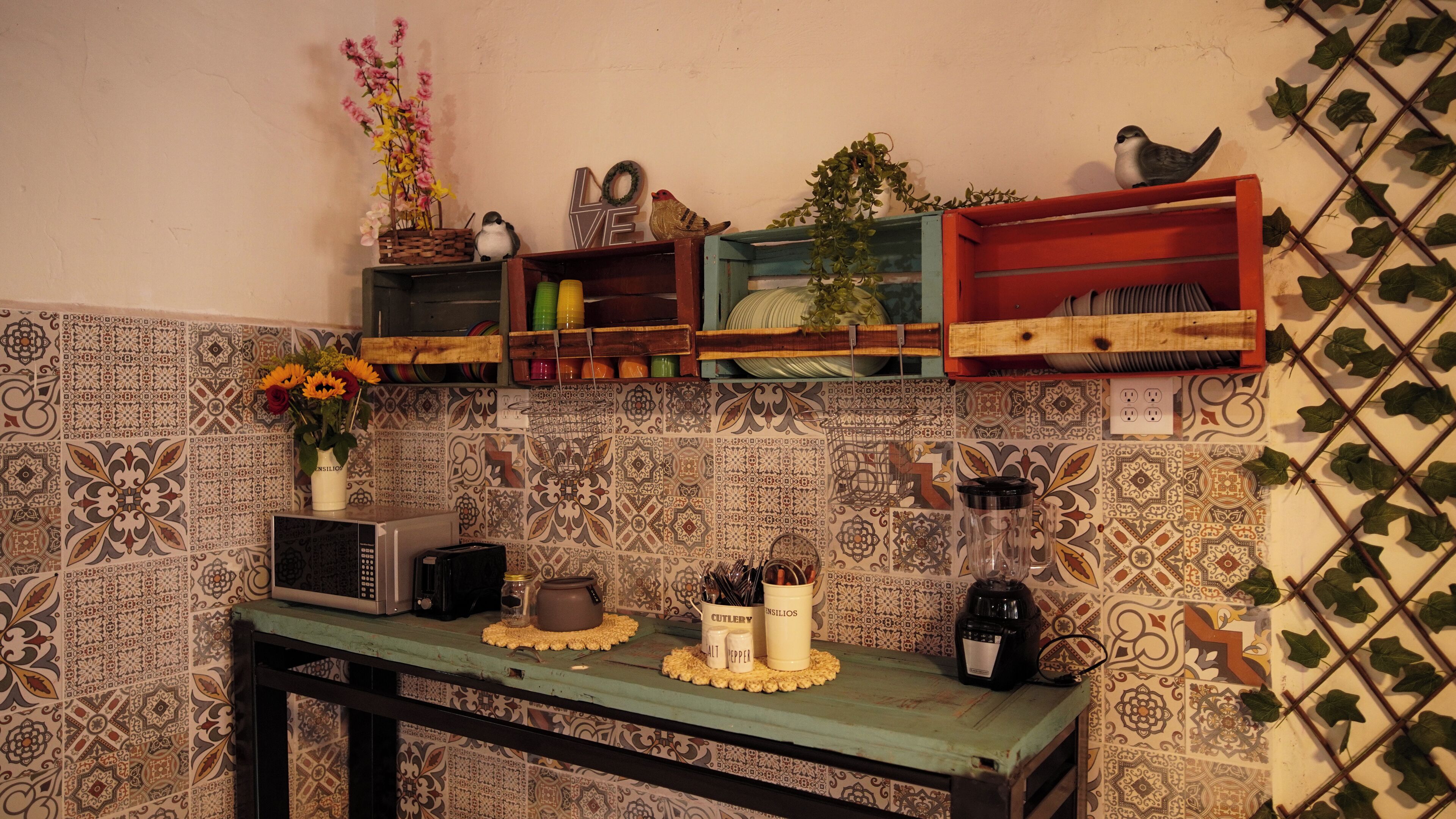 Uma cozinha com revestimento de azulejos, uma mesa de madeira com eletrodomésticos e prateleiras com diversos objetos.