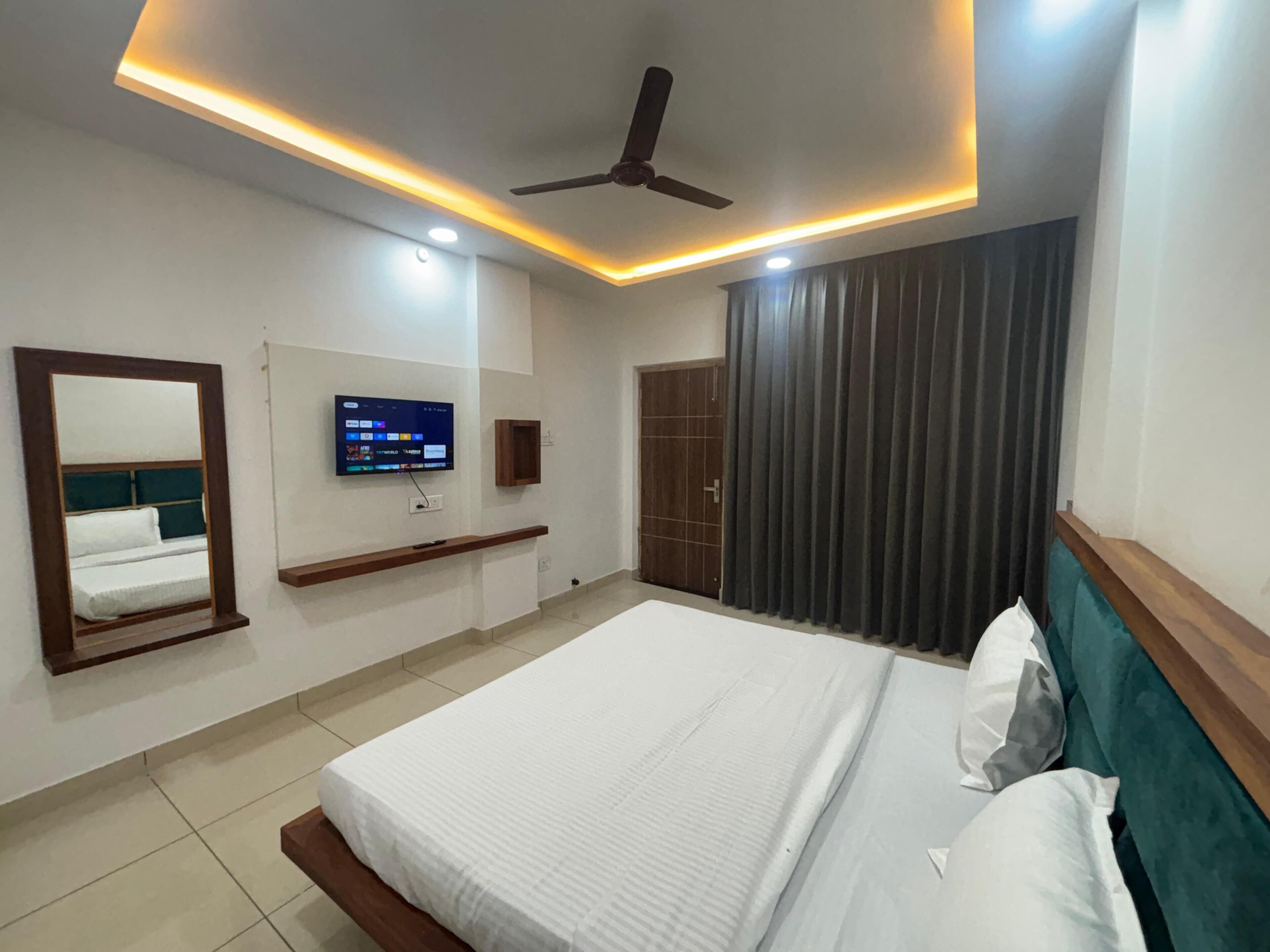 Quarto moderno com cama, ventoinha no teto, televisão e uma janela grande com cortinas.