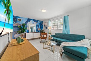 Uma sala de estar moderna com um sofá verde-água, uma mesa de centro de madeira, uma mesa de apoio com tampo de vidro, um espelho e uma parede com um mural tropical.