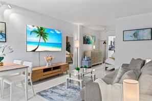 Uma sala de estar moderna com uma televisão de tela plana grande, uma área de jantar com mesa e cadeiras brancas, um sofá cinza e uma cozinha ao fundo.