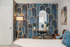 Quarto com cama, mesa de cabeceira, espelho, abajur e parede com padrão geométrico.