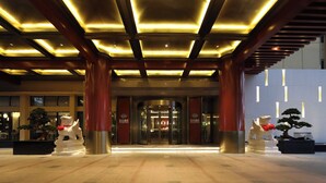 Entrada de hotel com design moderno, destacando uma fachada imponente com portas de vidro, teto iluminado e esculturas tradicionais.