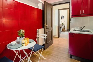 Uma cozinha compacta com armários vermelhos, bancada branca e um pequeno espaço de refeições com mesa e cadeiras.