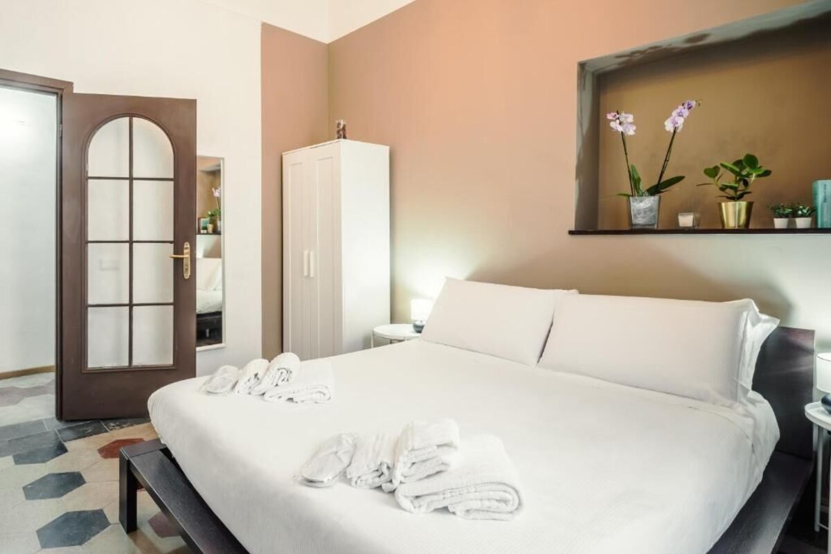 Quarto de hotel com cama, guarda-roupa, um banco, mesa de cabeceira e prateleira com plantas.