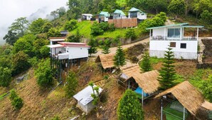 Uma povoação em encosta com vários edifícios, alguns com telhados de palha e outros com arquitetura moderna, cercada por vegetação exuberante.
