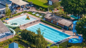 Uma grande piscina com escorrega e uma área de banheira de hidromassagem separada, rodeada por espreguiçadeiras e um pequeno edifício.