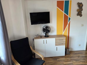 Um quarto com uma cadeira preta, um armário branco com gavetas, uma TV montada, um ventilador e uma lixeira.