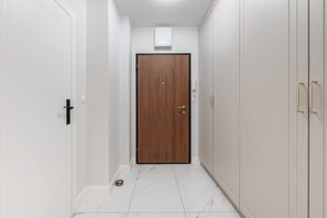 Um corredor com paredes brancas, uma porta de madeira no final e chão de azulejos brancos.