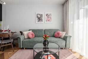 Uma sala de estar moderna com um sofá cinzento, duas almofadas cor-de-rosa, uma mesa de centro de vidro com revistas e um vaso com flores vermelhas.