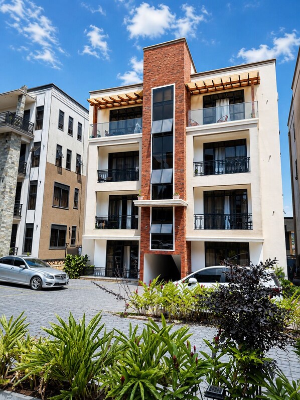 Um edifício residencial com vários andares, varandas, um destaque de tijolos no centro e uma entrada com estacionamento.