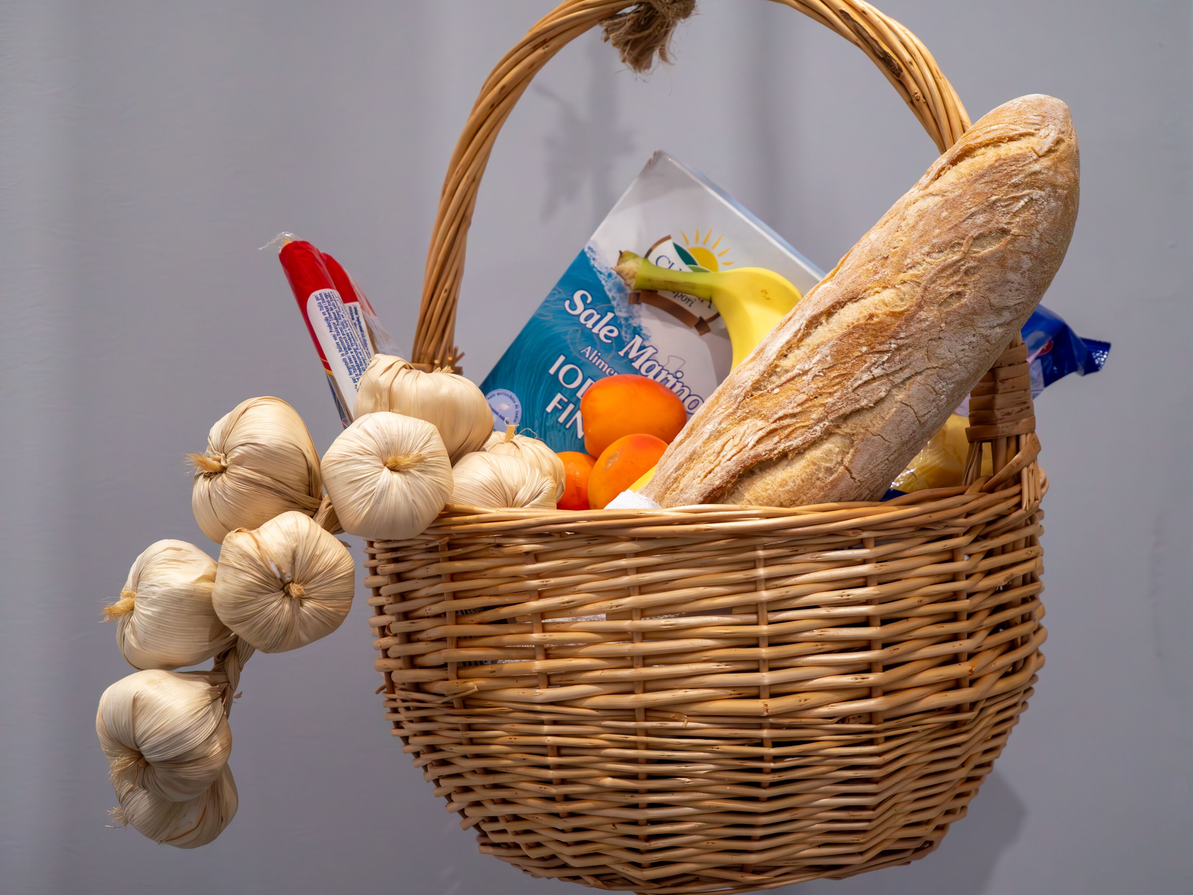 Uma cesta de vime com alho, uma baguete, laranjas e uma garrafa de azeite.