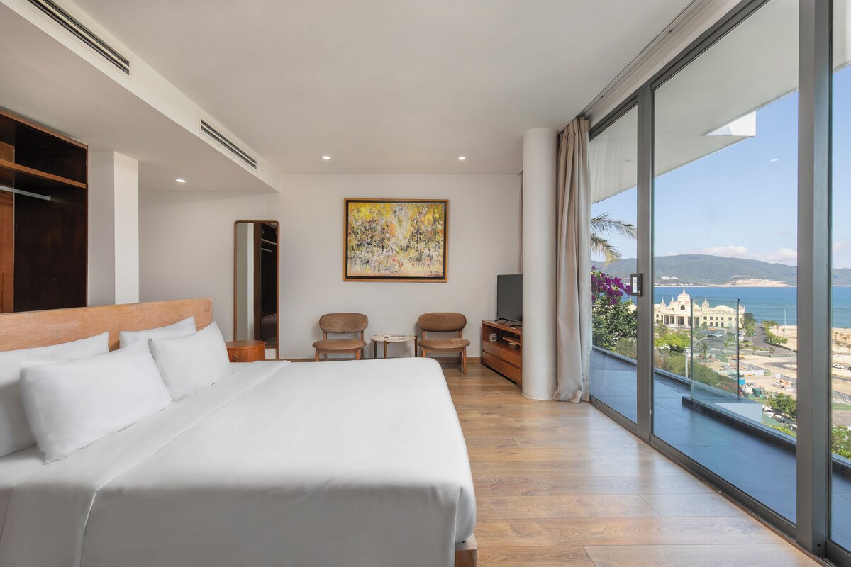 Quarto de hotel com uma cama grande, duas cadeiras, uma mesinha, um quadro na parede e vista para o mar e edifícios.