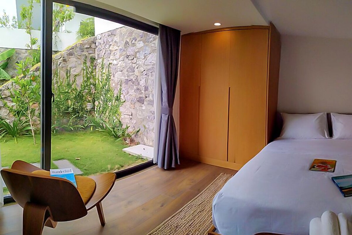 Quarto de hotel com cama, poltrona, guarda-roupa e vista para o exterior.