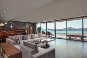 Ampla sala de estar com um sofá grande, uma mesa de jantar e um piano. Há uma janela grande que oferece vista para o mar e as colinas.